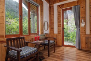 Interior de madeira com mesa e cadeiras, janela com vista para vegetação e porta com cortina.