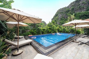 Piscina com espreguiçadeiras e guarda-sóis, cercada por vegetação exuberante e montanhas ao fundo.