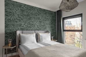 Quarto com cama grande, mesa de cabeceira, luminária e janela com vista para árvores.