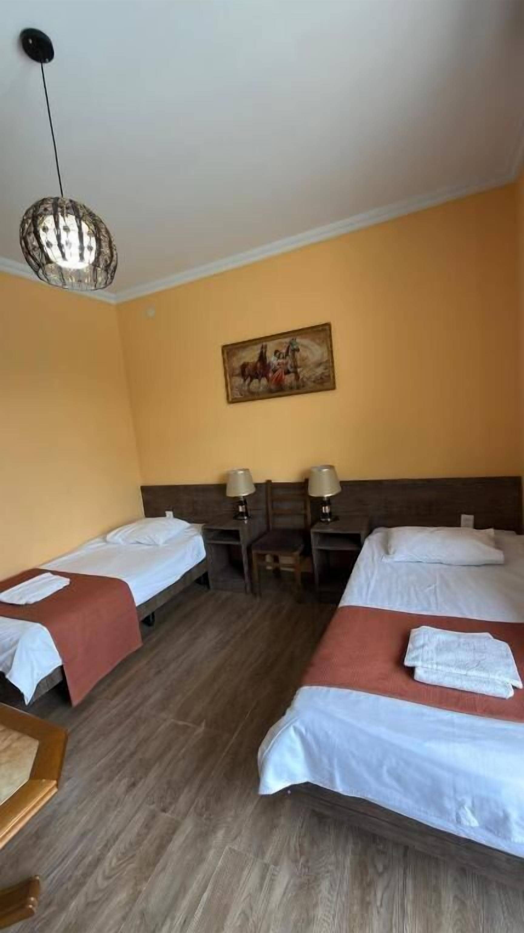 Quarto de hotel com duas camas de solteiro, piso de madeira, um quadro na parede e luminária pendente.
