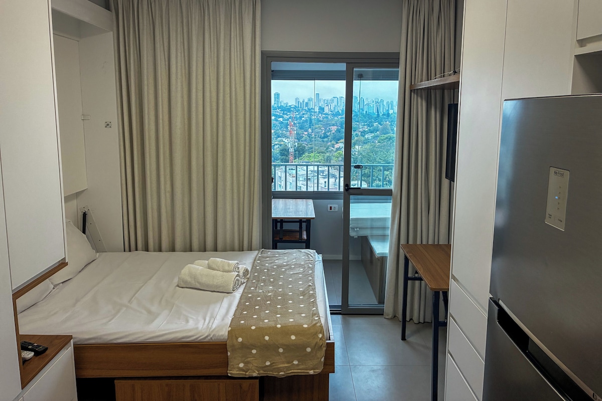 Quarto de hotel com cama, uma pequena mesa, geladeira e varanda com vista para a cidade.