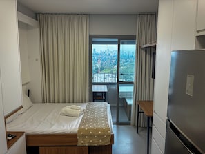 Quarto de hotel com cama, uma pequena mesa, geladeira e varanda com vista para a cidade.