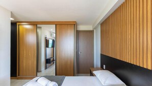 Quarto de hotel moderno com cama, painel de parede de madeira, guarda-roupa e banheiro com espelho e porta-toalhas.
