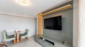 Uma sala de estar moderna com televisão de tela plana fixada em uma parede cinza, uma estante de madeira abaixo e um espaço de jantar com mesa e cadeiras.