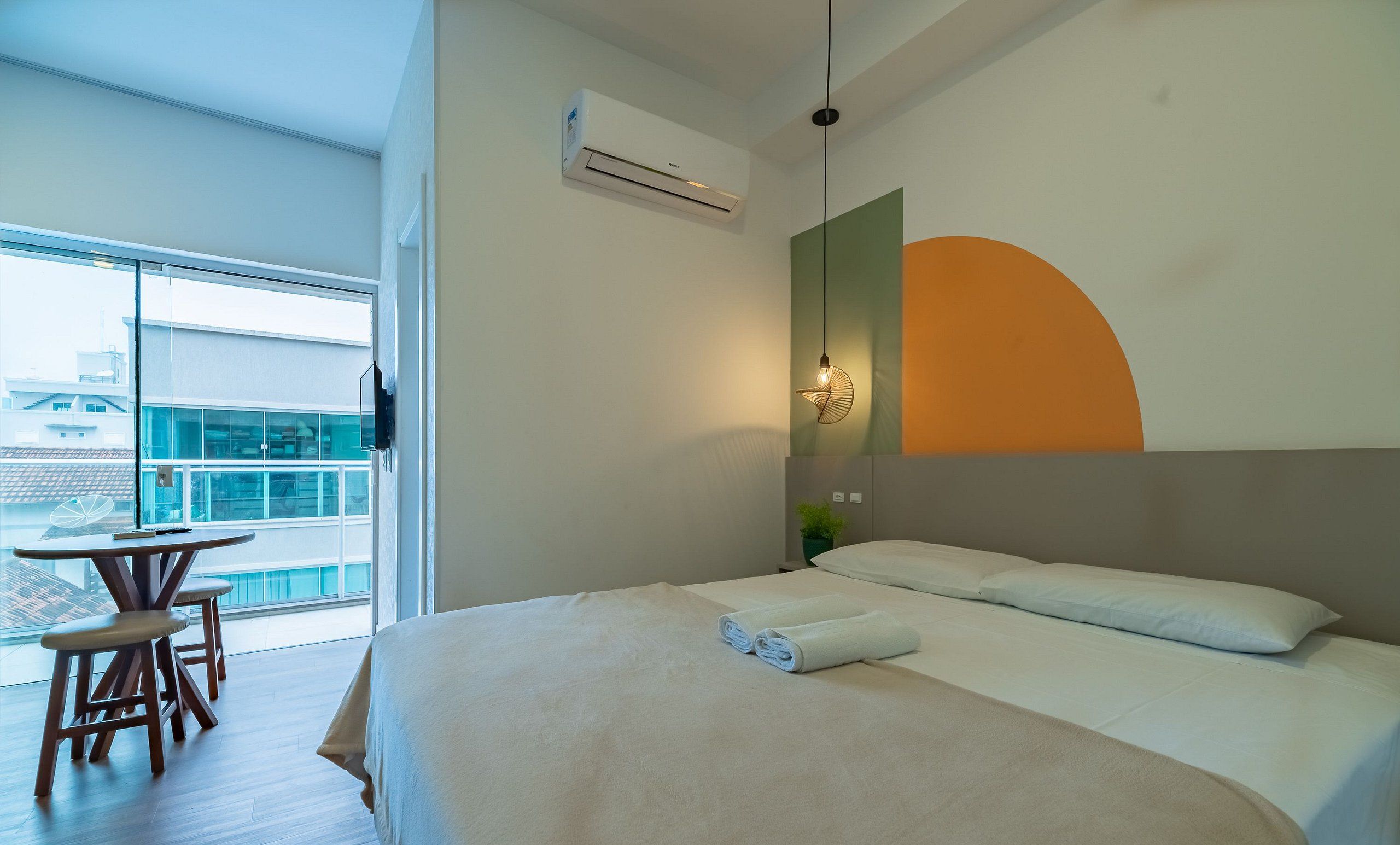 Quarto de hotel com uma cama, uma mesa e uma cadeira. Há uma janela com vista para prédios.