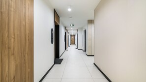 Um corredor comprido e estreito com portas em ambos os lados, piso de azulejos e teto com iluminação embutida.