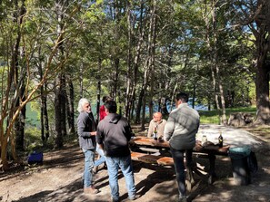 Um grupo de pessoas reunido em torno de uma mesa de piquenique em uma área arborizada, com um lago ao fundo.