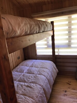 Uma cama beliche com colchão branco e cabeceira de madeira em quarto com paredes de madeira e janela com persianas.