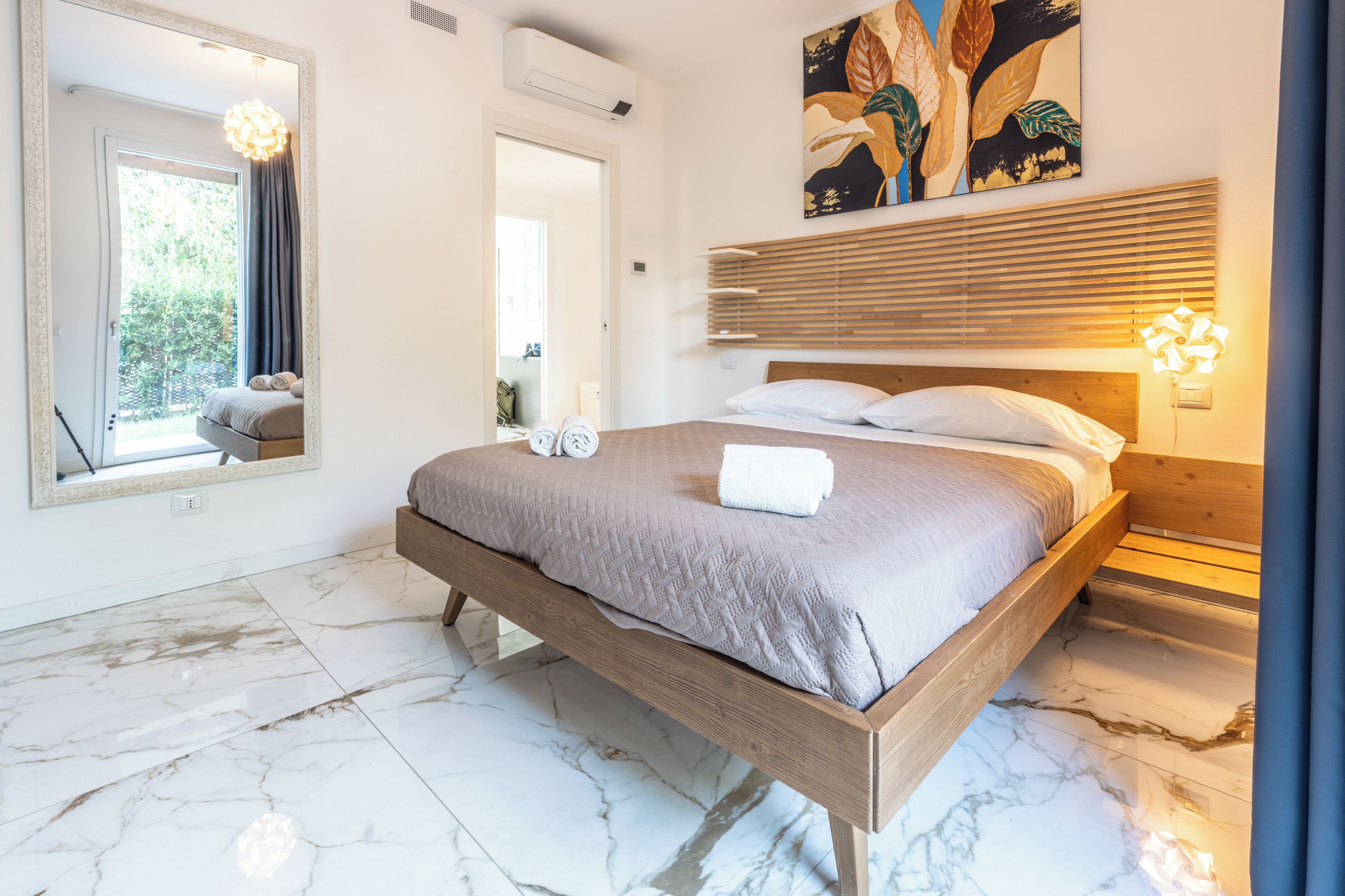 Quarto com cama de madeira, piso de mármore, um espelho grande e um quadro na parede.