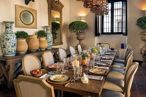Uma sala de jantar com uma mesa de madeira comprida posta para uma refeição, rodeada de cadeiras.