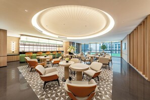 Um saguão de hotel moderno com área de estar central, balcão de recepção e grandes janelas.