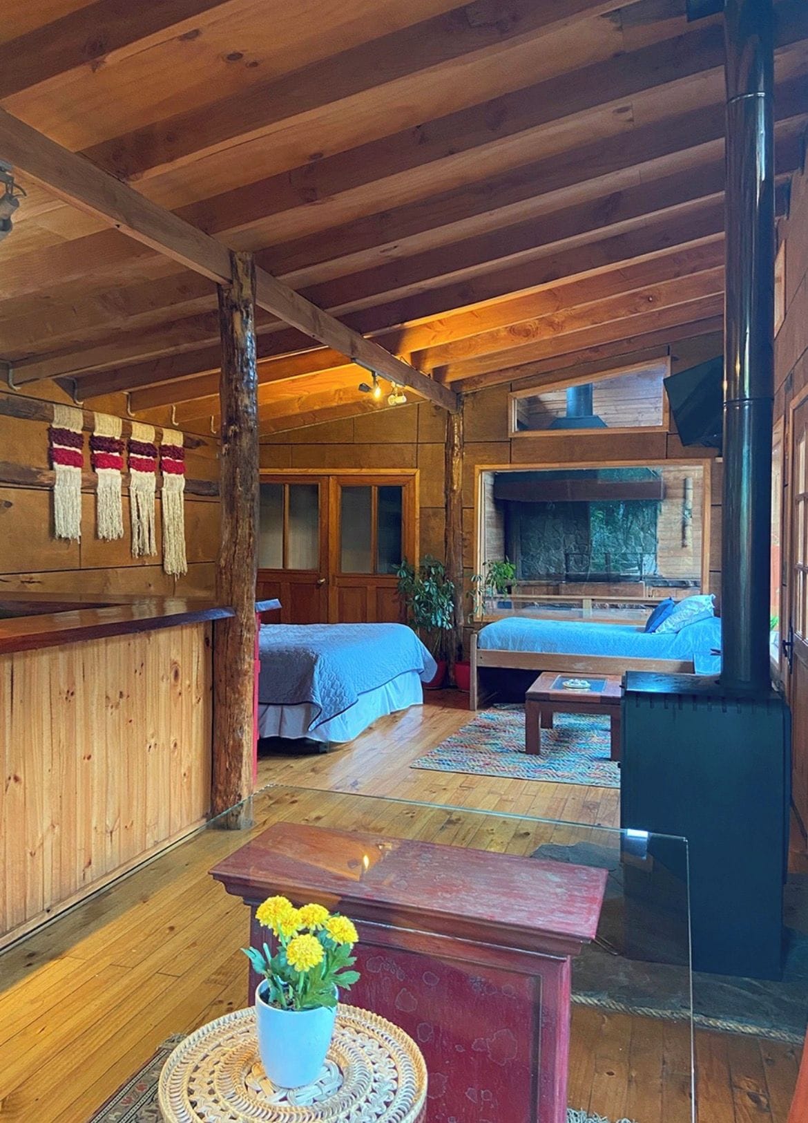 Interior de uma cabana de madeira com lareira, duas camas, um balcão de madeira e uma pequena mesa com uma planta.