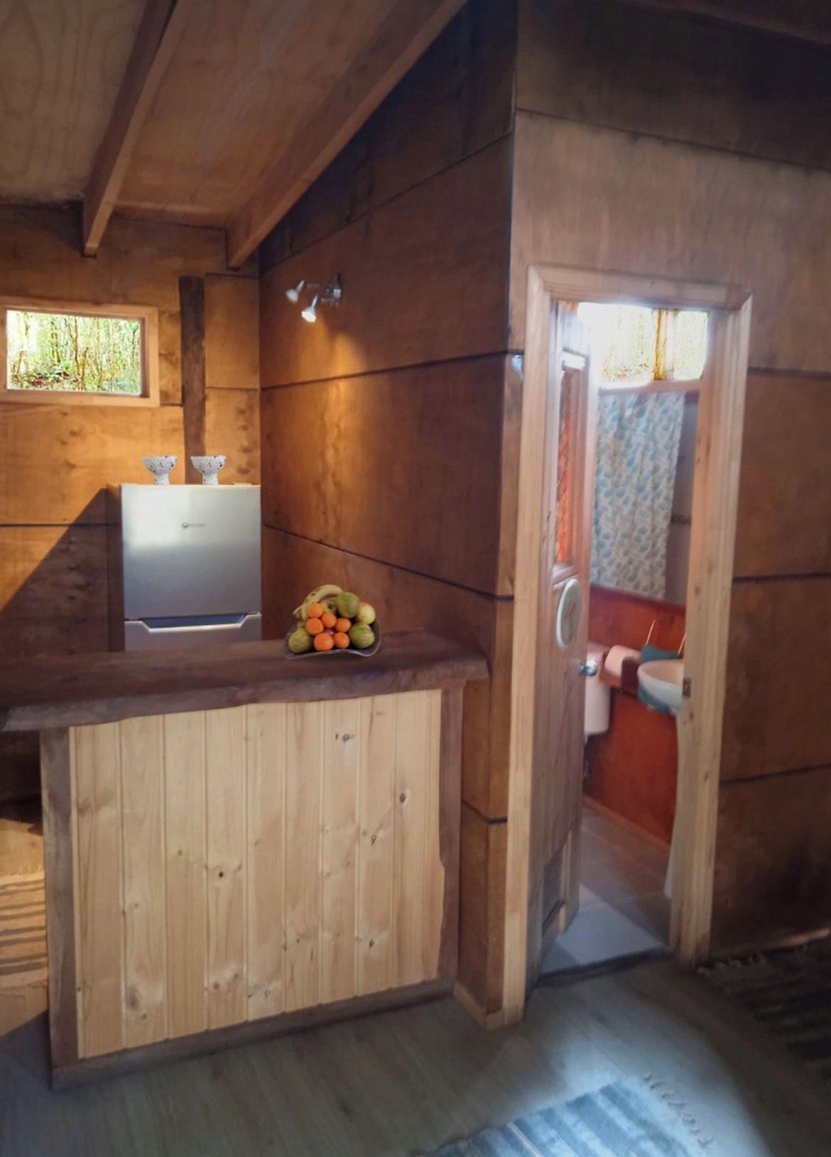 Interior de cabana de madeira, com bancada de cozinha, janela pequena e porta que dá para um banheiro.