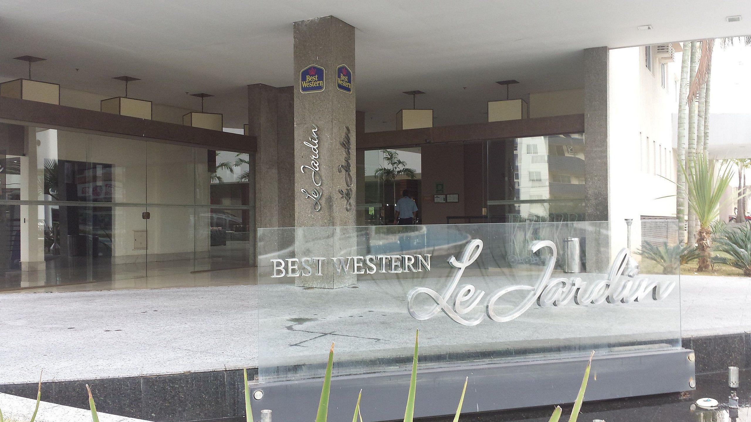 Entrada de hotel com fachada de vidro, placa com a inscrição 'BEST WESTERN Le Jardin' e uma pessoa visível no interior.