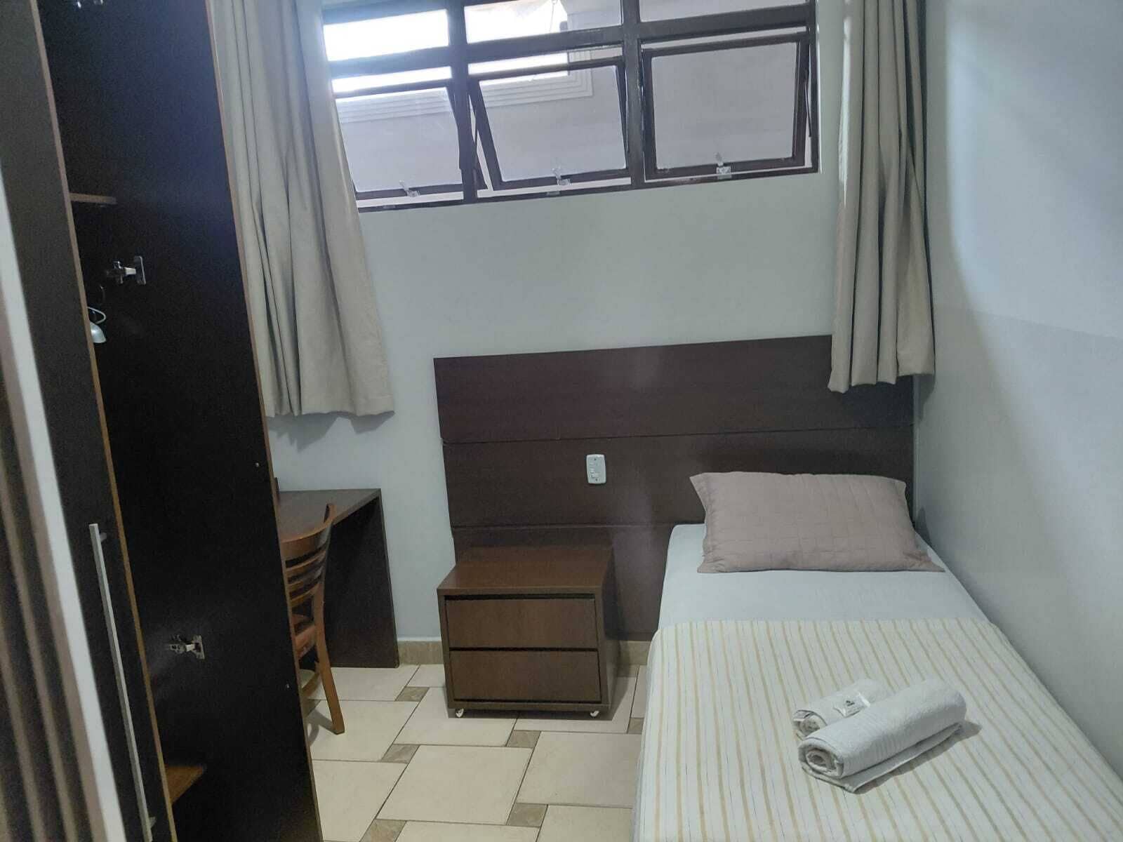 Uma cama de solteiro com colcha listrada, uma mesa de cabeceira de madeira, um pequeno armário de madeira e uma janela com cortinas.