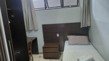 Uma cama de solteiro com colcha listrada, uma mesa de cabeceira de madeira, um pequeno armário de madeira e uma janela com cortinas.