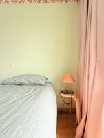 Uma cama de solteiro com colchão e travesseiro cinzas, uma pequena mesa de cabeceira com abajur e uma cortina à direita.