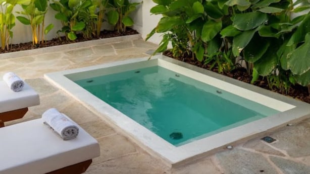 Uma banheira de hidromassagem retangular com interior azul claro, rodeada por soalho de pedra e plantas verdes exuberantes.
