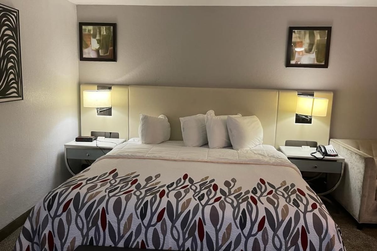 Quarto de hotel com uma cama grande, duas mesas de cabeceira, um criado-mudo e duas luminárias fixadas na parede.
