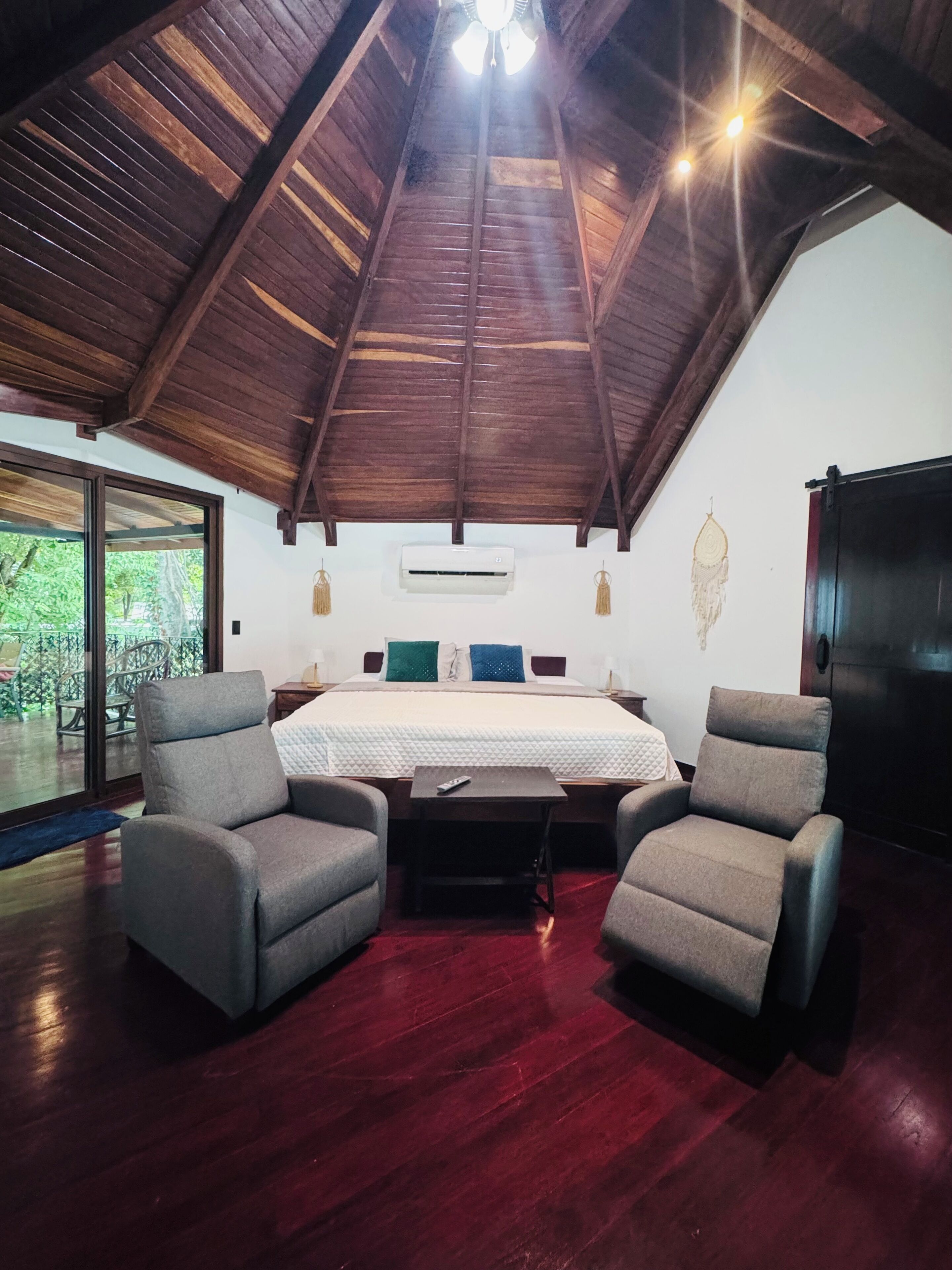 Quarto com teto de madeira, cama com almofadas, duas poltronas e uma janela grande.
