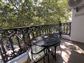 Varanda com mesa e cadeira de metal, cercada por grades pretas ornamentadas, e vista para árvores e um edifício ao fundo.