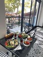 Mesa de jantar com diversos alimentos e bebidas, sobre um piso estampado, com vista para uma varanda e área de estar ao ar livre.