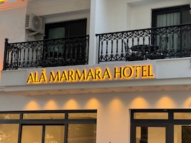Exterior do Hotel Alá Marmara com um letreiro iluminado, uma varanda com grades pretas ornamentadas e um grande aparelho de ar condicionado fixado no prédio.