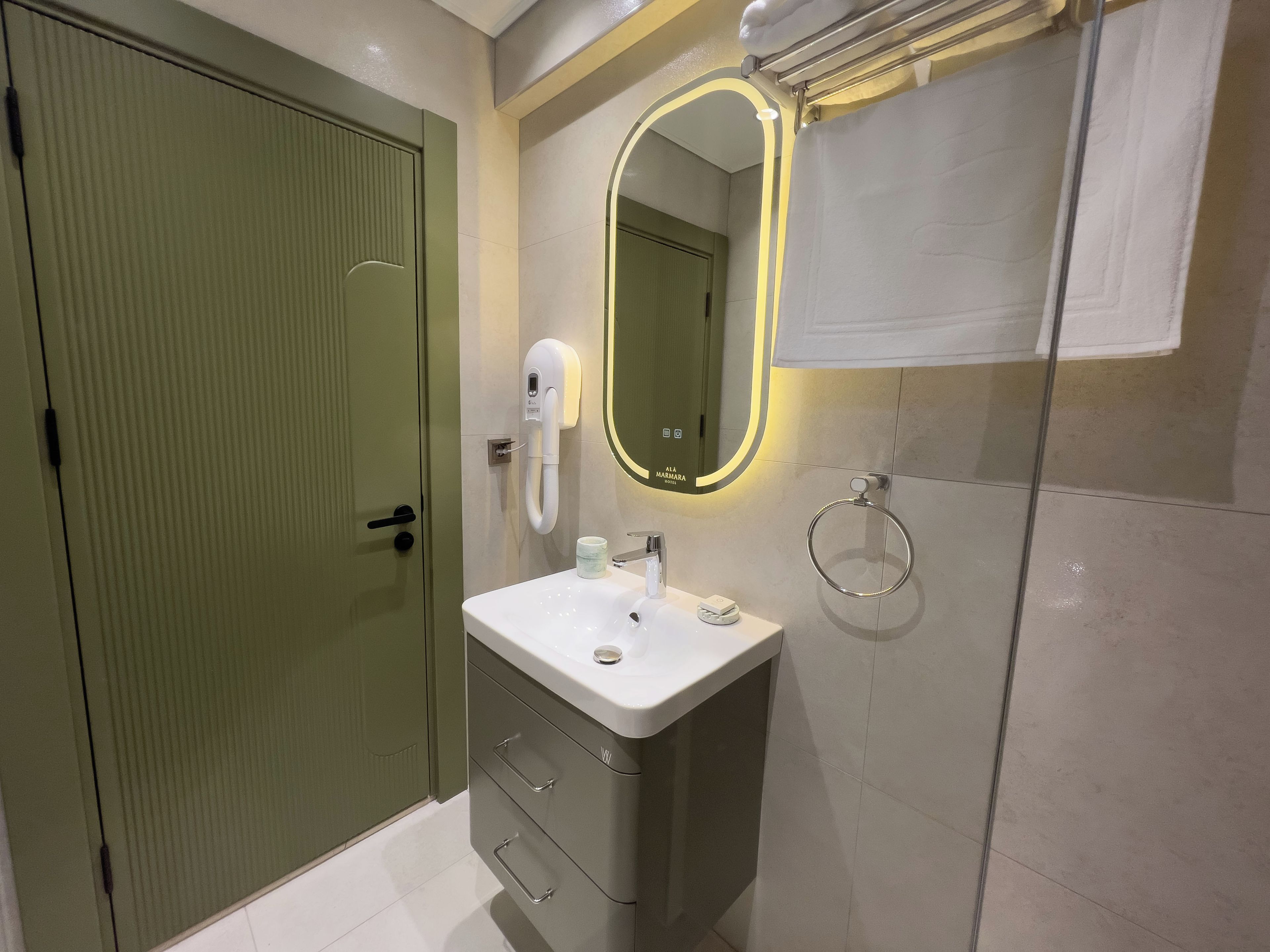 Casa de banho com porta verde, pia branca, espelho iluminado e suporte para toalha.