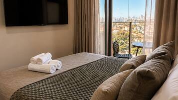 Quarto de hotel com cama grande, televisão de tela plana na parede, varanda com vista para a cidade e mesa com cadeira na área externa.