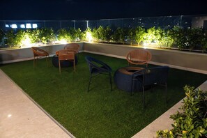 Terraço no telhado com grama sintética, cadeiras de vime e plantas em vasos.