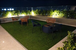 Terraço no telhado com grama sintética, cadeiras de vime e plantas em vasos.