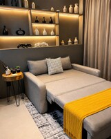 Quarto moderno com sofá-cama, mesinha lateral, manta amarela e prateleira na parede com objetos decorativos.