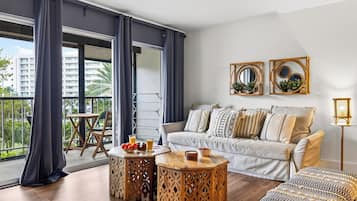 Uma sala de estar moderna com um sofá, duas mesinhas laterais e uma varanda com vista para um edifício e palmeiras.