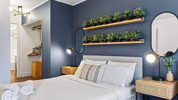 Quarto moderno com cama, mesas de cabeceira, espelho e prateleiras fixas com plantas.