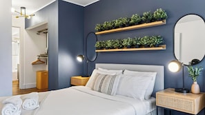 Quarto moderno com cama, mesas de cabeceira, espelho e prateleiras fixas com plantas.