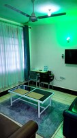Uma sala de estar com ventilador de teto, TV de tela plana na parede, mesa de centro com papéis, uma cadeira e um sofá.
