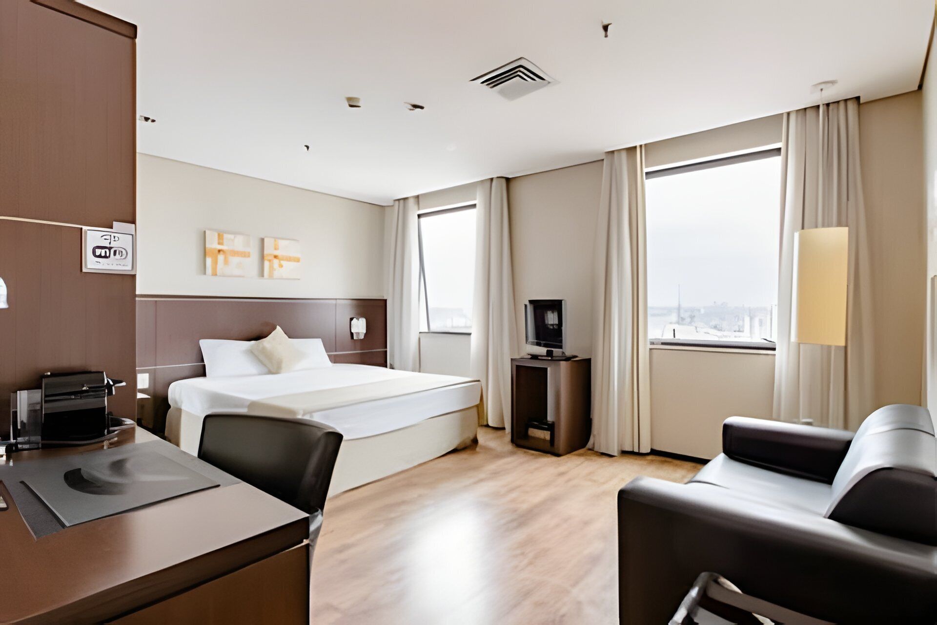 Quarto de hotel com cama, escrivaninha com impressora, televisão e vista para a cidade através de amplas janelas.