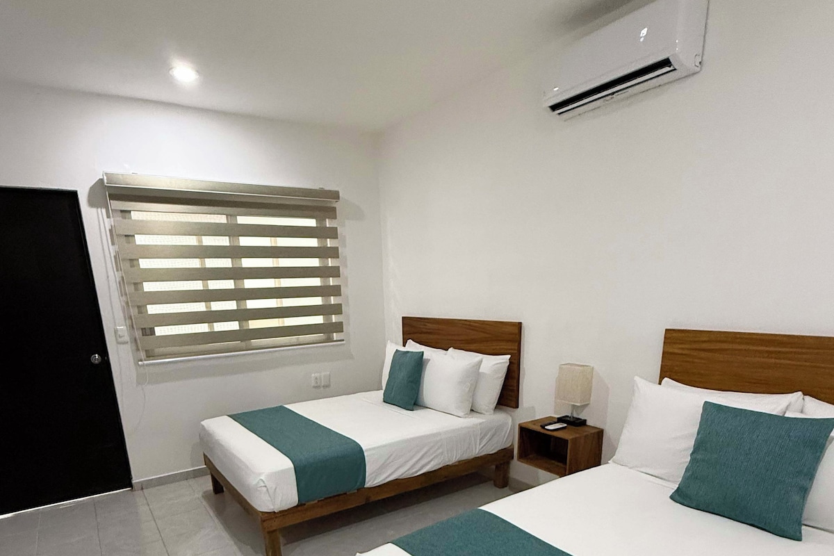 Quarto de hotel com duas camas, janela com persianas, ar condicionado e piso de azulejos.