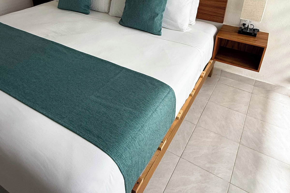 Cama com cabeceira de madeira, roupa de cama branca e almofadas azuis-esverdeadas.