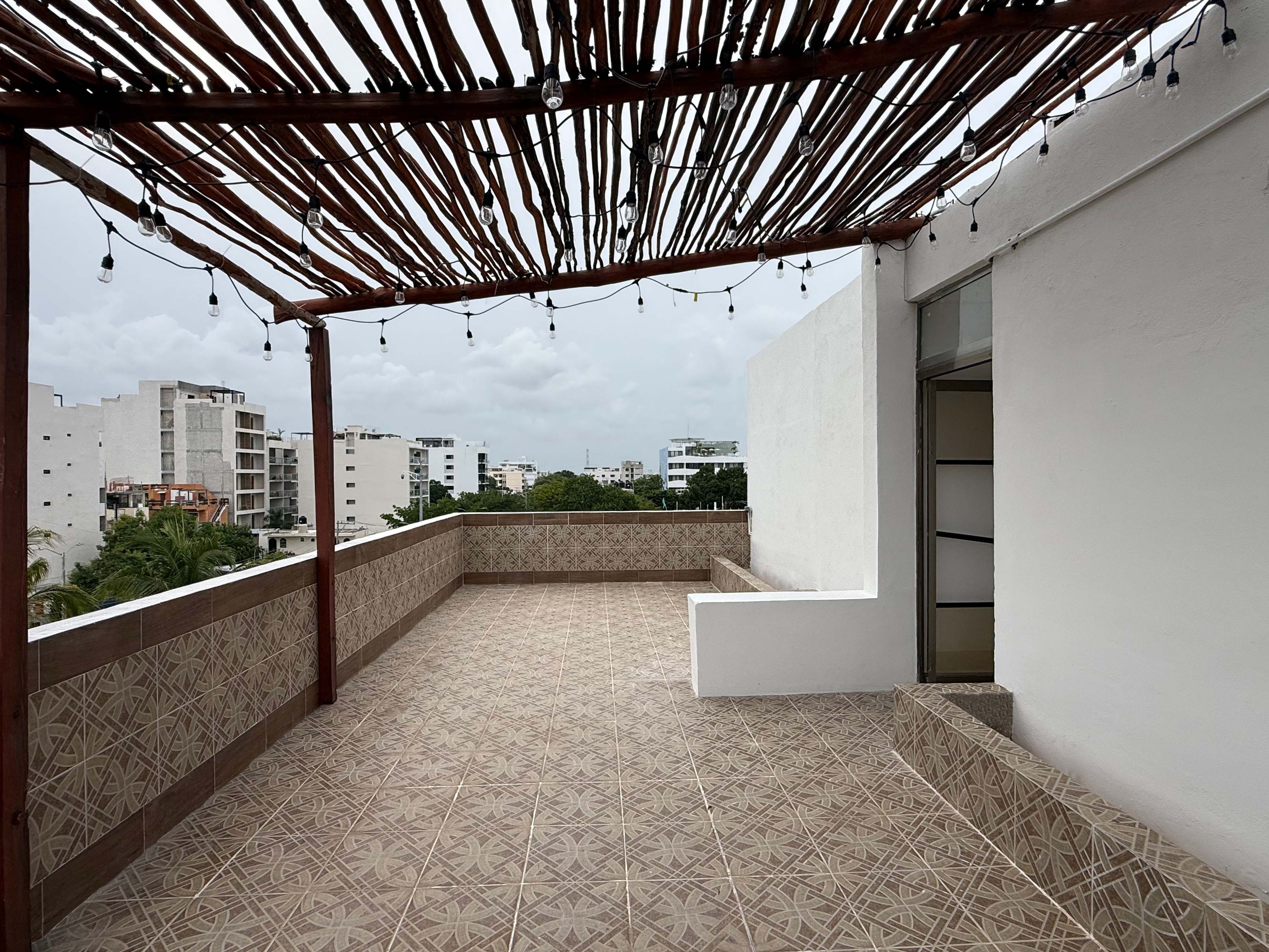 Terraço no telhado com ladrilhos estampados, pérgola de madeira com luzes de cordão e vista dos edifícios ao redor.