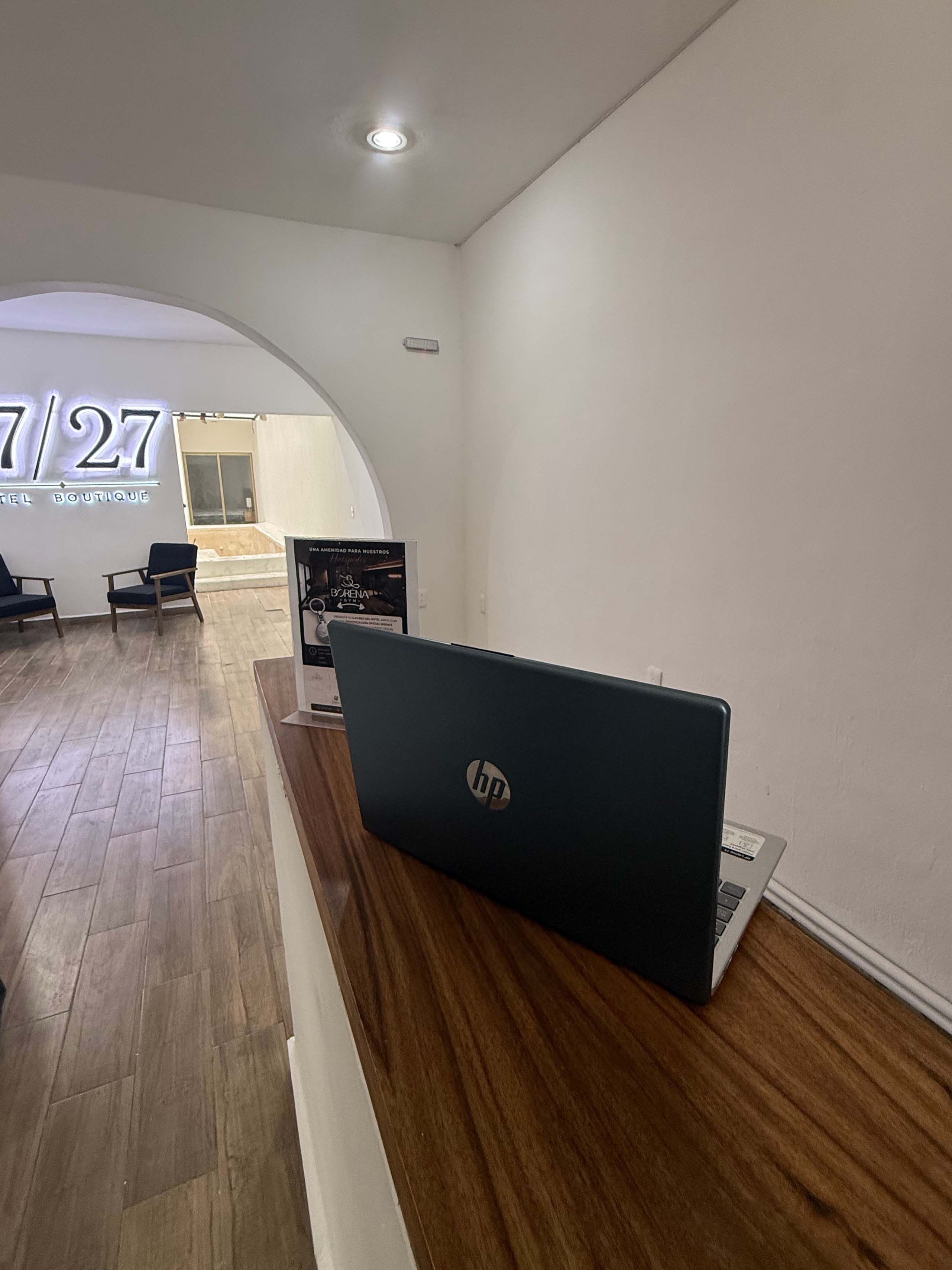 Uma área de recepção de hotel com um balcão de madeira, um laptop HP e uma placa com a inscrição '7/27 Boutique Hotel'.
