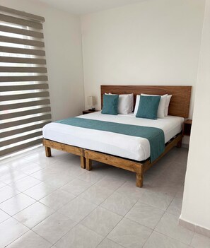 Cama bem arrumada com estrutura de madeira e roupa de cama branca, mesa de cabeceira com abajur e janela com persianas horizontais.