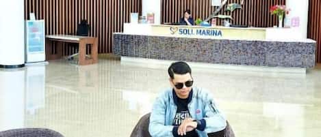 Um homem sentado em uma cadeira com um laptop em uma área de lobby com uma mesa de recepção identificada como 'SOLL MARINA'.