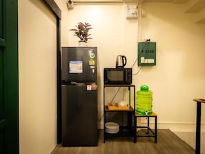 Uma cozinha compacta com geladeira preta, micro-ondas, prateleira pequena com louças, chaleira e dispenser de água verde.