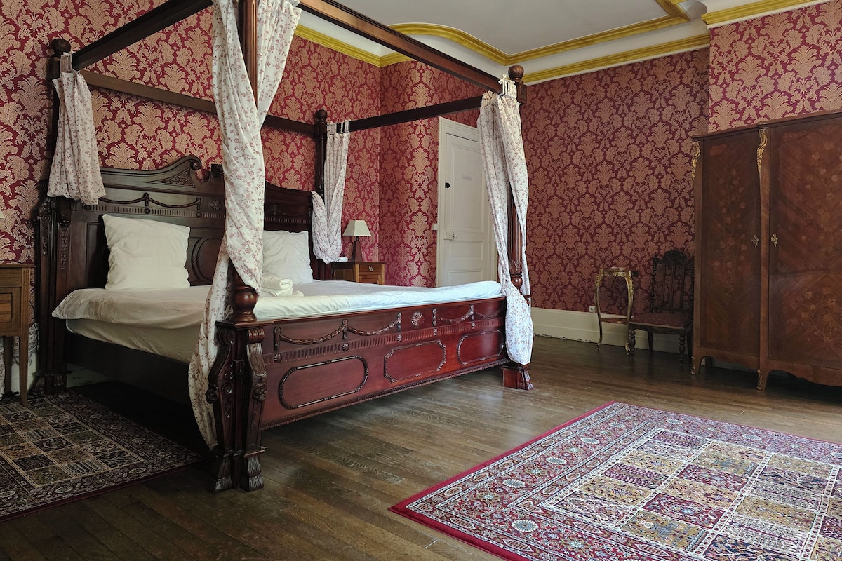 Uma cama com dossel e cortina, um guarda-roupa de madeira e um tapete estampado em um quarto com papel de parede decorado.