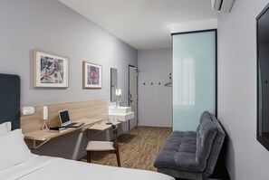 Quarto de hotel com cama, escrivaninha, cadeira e sofá cinzento. Há duas obras de arte emolduradas na parede, um espelho e uma área de chuveiro.