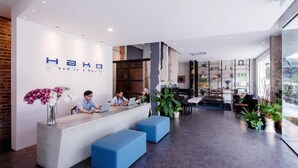 Uma área de recepção de hotel moderna, com balcão de atendimento, dois funcionários e um espaço de estar com almofadas azuis.