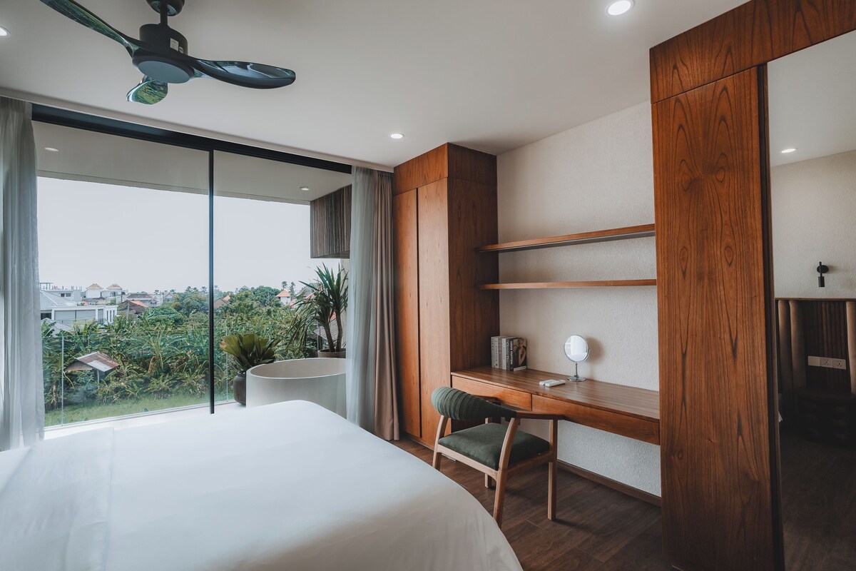 Quarto moderno com cama grande, escrivaninha com prateleiras, cadeira e vista para o verde lá fora.