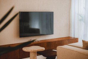 Uma sala de estar moderna com uma TV de tela plana fixada na parede, um armário de madeira embaixo, um banquinho redondo pequeno e um sofá bege.