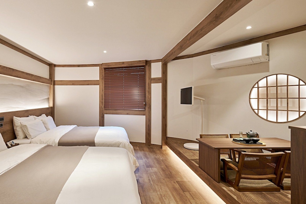 Quarto de hotel com duas camas, uma mesa de jantar com cadeiras, uma televisão e um aparelho de ar condicionado.